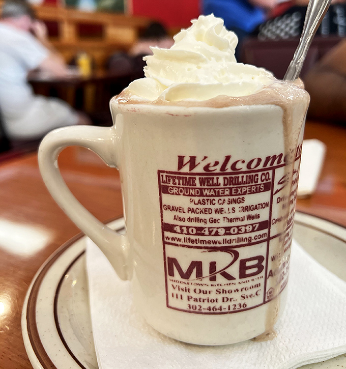 Hot chocolate that understands whipped cream isn't a topping&mdash;it's a lifestyle choice. The mug advertising local businesses is peak small-town charm.