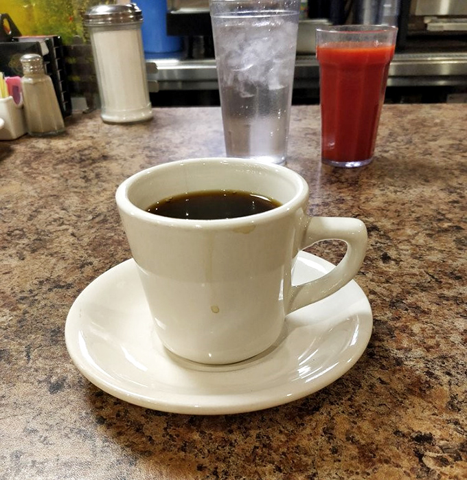The diner coffee trinity: sturdy white mug, saucer that's seen it all, and tomato juice nearby for those who believe in breakfast balance.