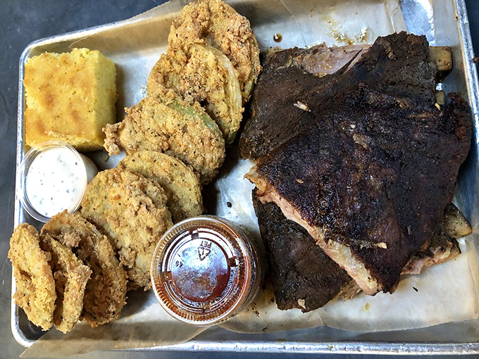 A tray of ribs with the perfect bark, flanked by golden cornbread and fried green tomatoes &ndash; the holy trinity of Southern comfort on a metal tray.