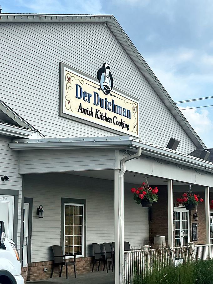 The unassuming exterior of Der Dutchman belies the culinary treasures within. Simple white siding, hanging flower baskets, and that iconic sign promising authentic Amish cooking.