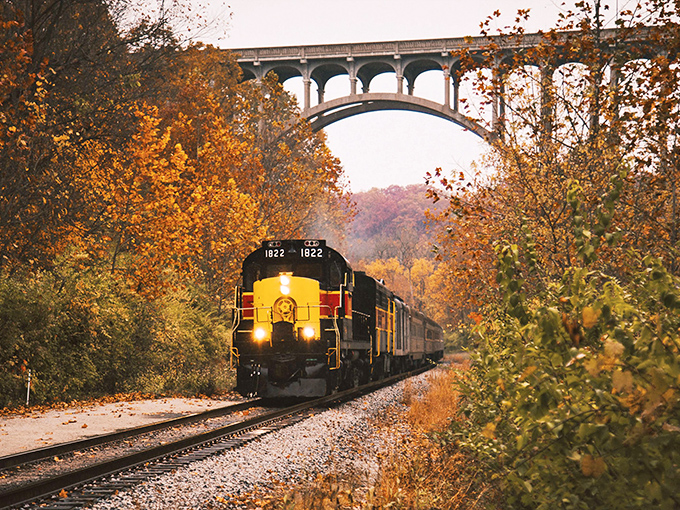 Fall's fiery canvas unfolds as the iconic yellow and red locomotive glides beneath a stone arch bridge, creating a scene straight from a vintage postcard.