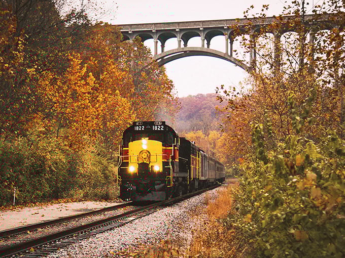 Fall's fiery canvas unfolds as the iconic yellow and red locomotive glides beneath a stone arch bridge, creating a scene straight from a vintage postcard.