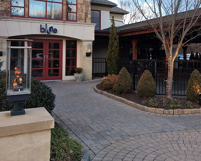 The entrance to Blue Grillhouse welcomes you with warm stone, distinctive blue signage, and the promise of culinary adventures waiting just beyond those red doors.