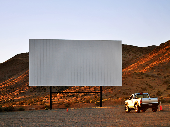 Cars line up at dusk facing the massive white screen, where cinematic magic awaits under the vast Mojave sky.