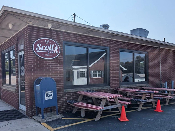 The brick-faced charm of Scott's Diner beckons from the roadside, promising comfort food salvation with those patriotic picnic tables standing at attention.