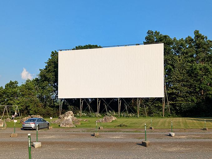 The massive white screen stands ready against the blue sky, with cars already claiming prime viewing spots. Movie magic awaits as the sun begins its descent.