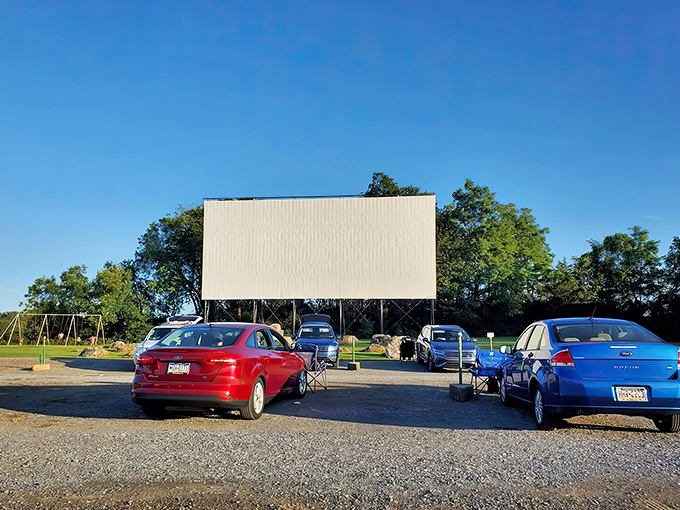 The massive white screen stands ready against the blue sky, with cars already claiming prime viewing spots. Movie magic awaits as the sun begins its descent.
