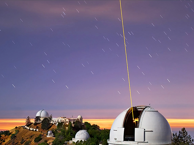 Starlight magic at its finest! The observatory's iconic white domes stand sentinel against a twilight sky, with a laser beam piercing the heavens like nature's own exclamation point.