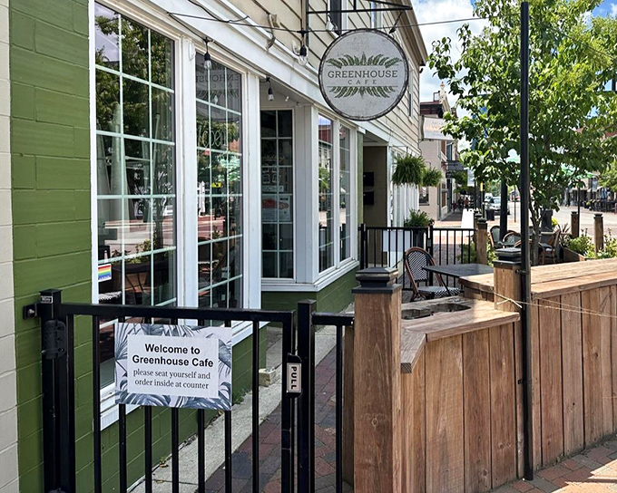 The Greenhouse Cafe welcomes you with its charming green exterior, classic white-framed windows, and a cozy outdoor seating area framed by natural wood&mdash;inviting you to come in, relax, and enjoy.