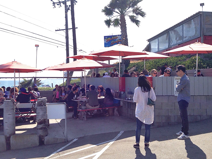 The outdoor dining area buzzes with happy eaters under cheerful orange umbrellas. Ocean views come standard with every meal.