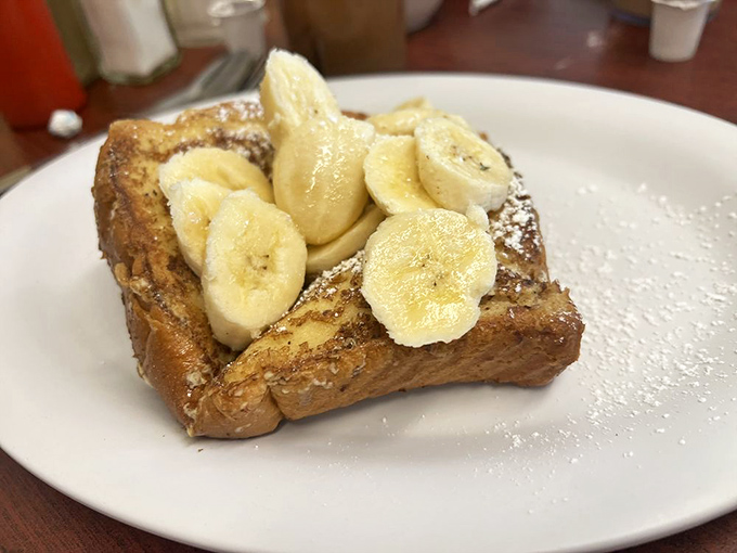 French toast that's achieved breakfast nirvana—crisp edges, custardy center, topped with fresh bananas and a dusting of powdered sugar that looks like morning snowfall.