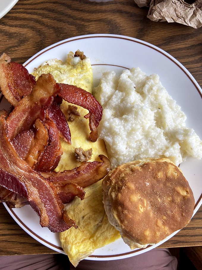 The breakfast trinity: crispy bacon, fluffy eggs, and grits with a golden biscuit standing guard. This plate doesn't need a prayer&mdash;it is the blessing.