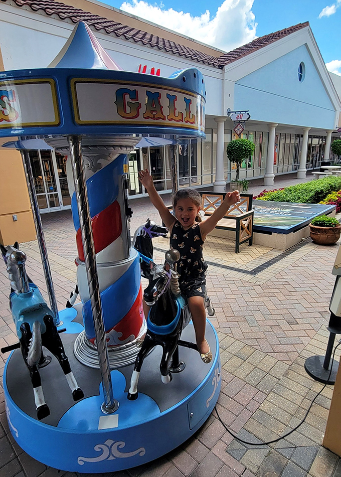 The universal expression of outlet mall victory: arms raised in triumph atop a carousel horse, shopping bags safely stashed nearby.