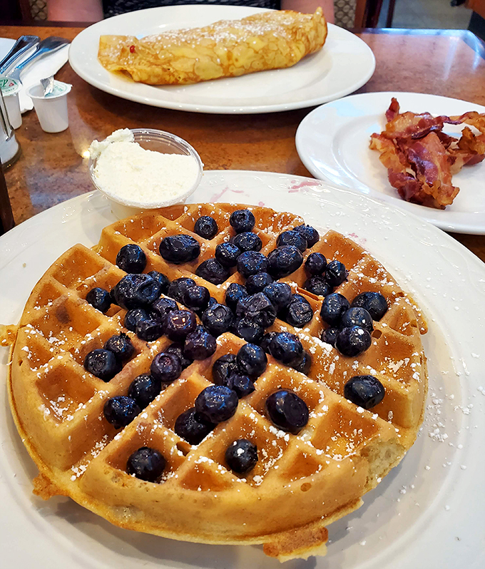 A waffle that's become a blueberry landing pad. The bacon in the background is just waiting for its maple syrup crossover event.
