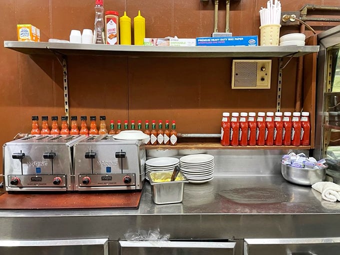 The condiment lineup stands ready for duty like tiny soldiers of flavor. Hot sauce bottles gleam under fluorescent lights, promising heat for the brave.