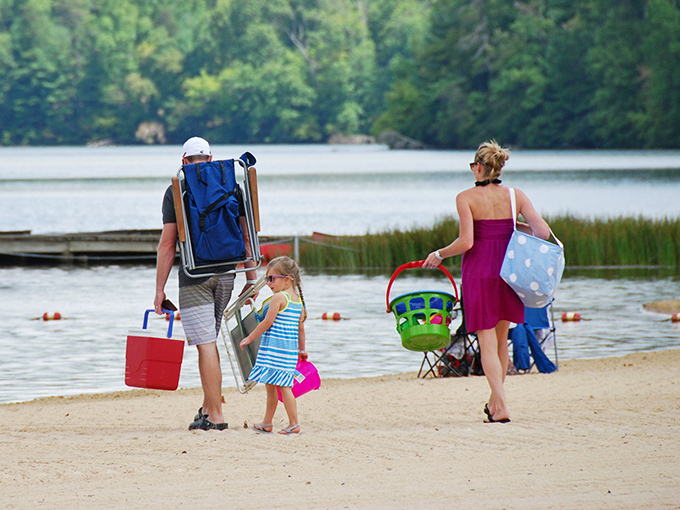 Families trek across golden sand carrying the universal symbols of lake day success: coolers, chairs, and excessive amounts of beach toys.