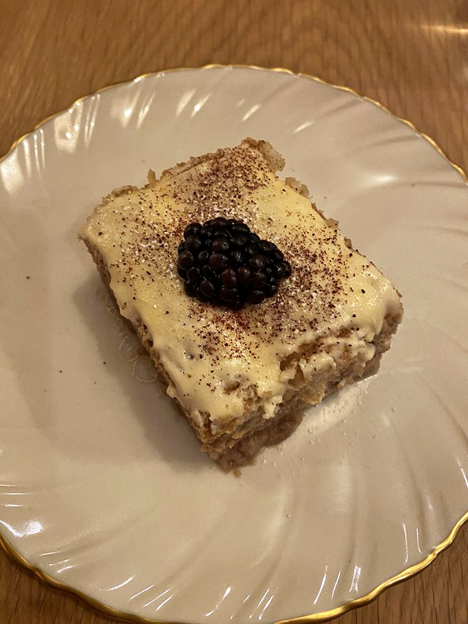 A square of tiramisu with a single blackberry guardian—coffee-soaked layers promising the perfect bittersweet finale.