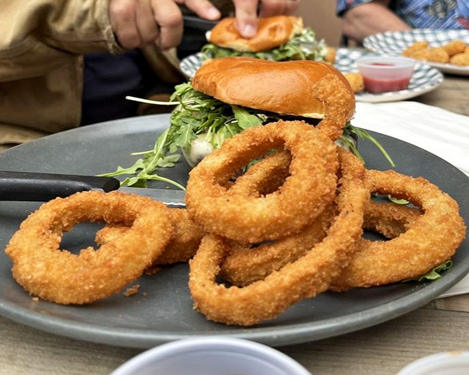 Onion rings stacked like delicious golden halos, proving that sometimes the side dish can steal the spotlight. That burger in the background isn't even mad about it.