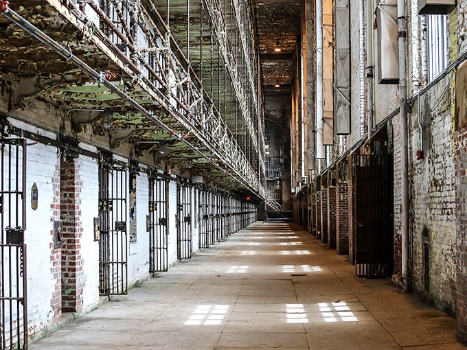 The corridor of confinement stretches toward infinity. Sunlight creates a haunting pattern of bars across the floor where countless inmates once walked.