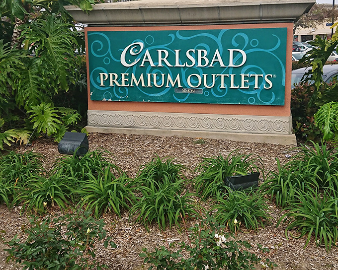 The entrance sign, surrounded by lush landscaping, promises retail paradise ahead. Like the pearly gates, but for shopping enthusiasts with champagne taste on a beer budget.