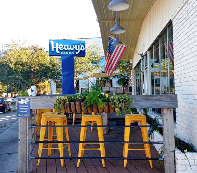 The outdoor seating area says "stay awhile" with its bright yellow stools and lush greenery framing that iconic blue sign.