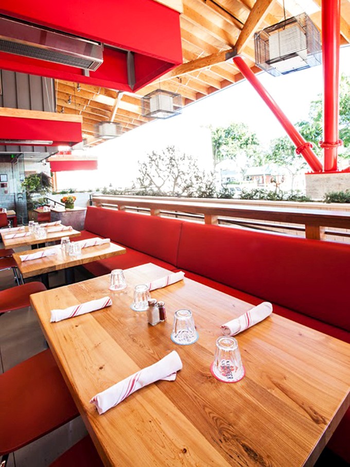The patio seating offers a breezy California dining experience, where the red accents pop against natural wood in a space that feels both indoors and out.