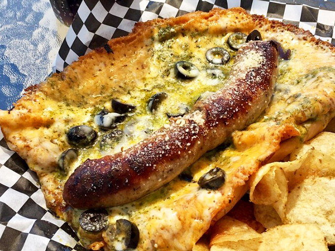 The Pesto Brat redefines sausage expectations &ndash; nestled in a golden cheese blanket with olives standing guard over this flavor masterpiece.