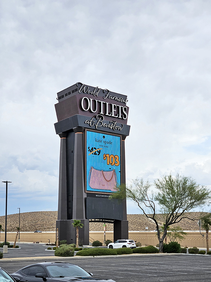 The towering sign stands as a retail lighthouse, guiding weary travelers toward discounted Kate Spade and away from full-price department stores.