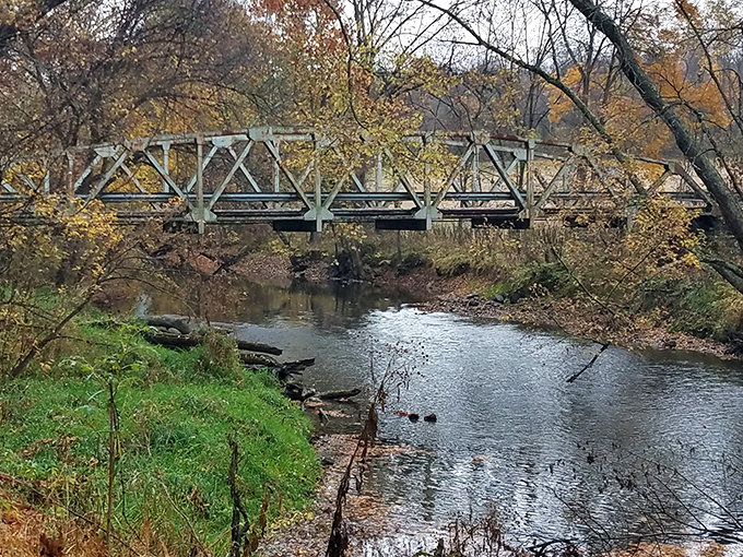 Nature's own gallery exhibition! This metal bridge spans the gentle Mohican River, framed by autumn foliage that would make Bob Ross reach for his palette.