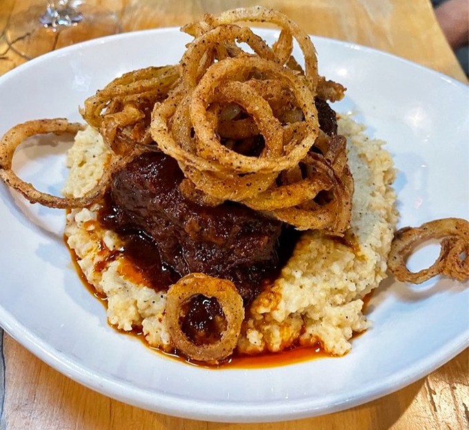 Behold the star attraction: short ribs that surrender to your fork, crowned with crispy onion rings atop a bed of creamy grits.