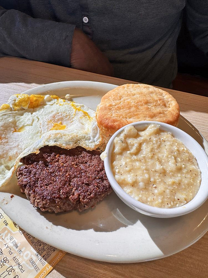 Breakfast perfection on a plate: a golden-yolked egg, seasoned burger patty, fluffy biscuit, and creamy grits. This is why alarm clocks were invented.