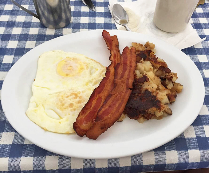 Breakfast perfection doesn't need fancy plating &ndash; just a sunny-side up egg, crispy bacon, and home fries on a blue-checked tablecloth.
