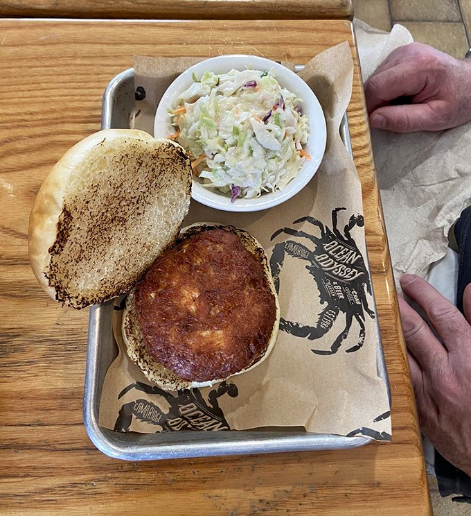 Behold seafood perfection: a golden-brown crab cake sandwich with minimal filler, maximum crab, served with creamy coleslaw on a metal tray lined with branded paper.