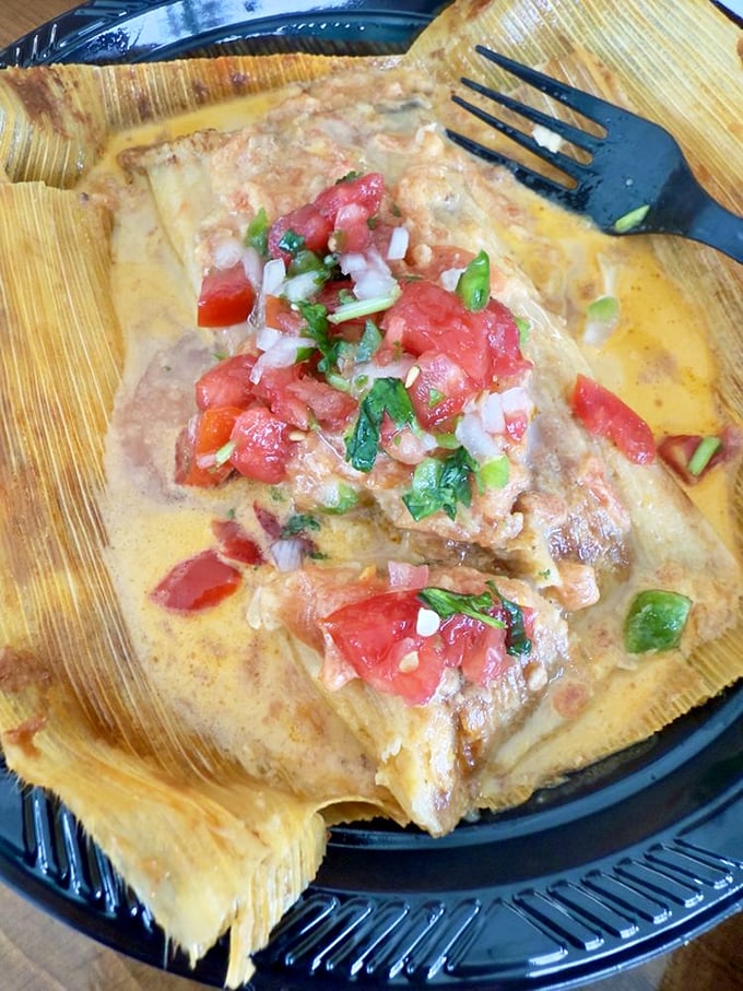 Behold the tamale in its corn husk throne! Fresh pico de gallo crowns this masa masterpiece like jewels on culinary royalty.