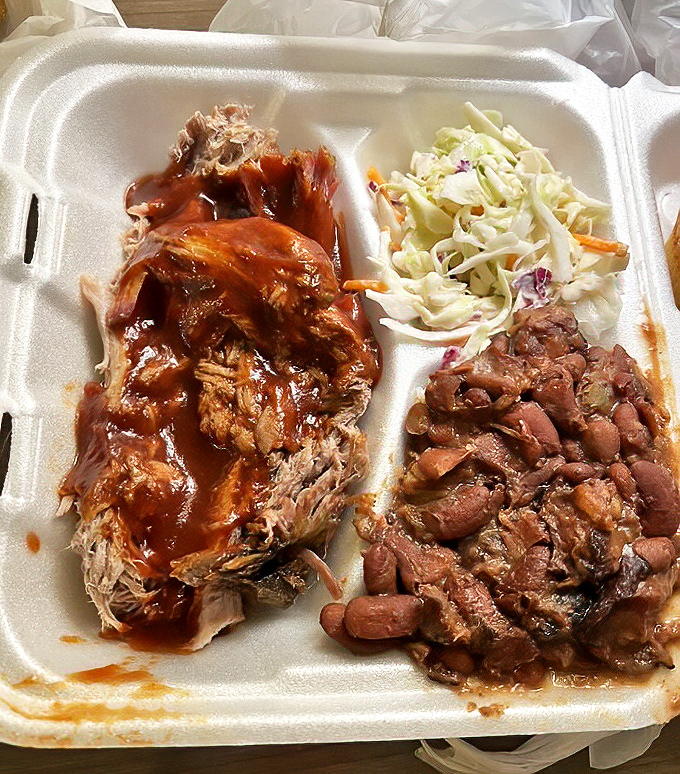 Pulled pork glistening with sauce alongside creamy slaw and beans. This styrofoam container holds more genuine happiness than most luxury restaurants could ever deliver.