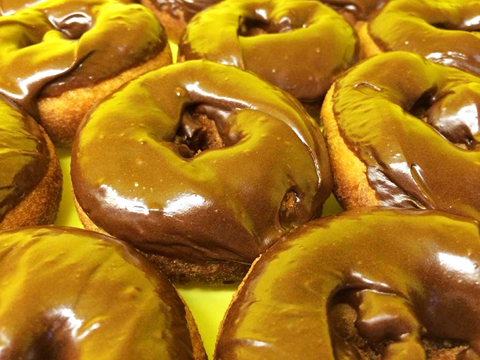 These chocolate-glazed beauties glisten with promise, their perfect sheen reflecting decades of donut-making expertise that no franchise could ever replicate.