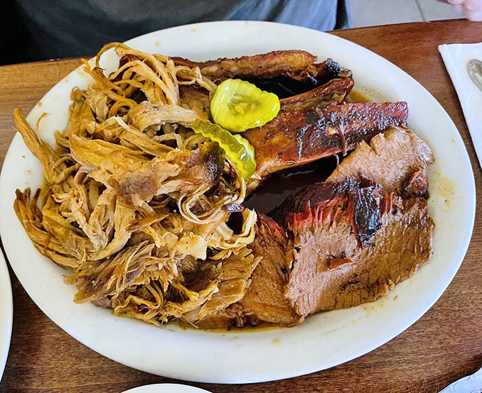 Behold the holy trinity of BBQ&mdash;brisket, pulled pork, and ribs sharing a plate in perfect harmony. Those pickle chips aren't just garnish; they're necessary acid relief.