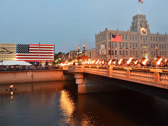 Evening lights and community pride shine bright in Sharon, Pennsylvania, as torches line the bridge and American flags wave proudly over this charming riverside town.