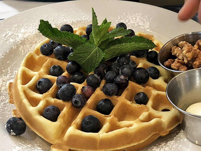 A Belgian waffle that's dressed for success with fresh blueberries, a mint garnish, and two sidecars of deliciousness. Syrup application is your sacred responsibility.