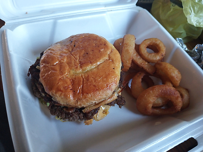 A classic diner burger nestled beside crispy onion rings in a takeout container &ndash; proof that simple food done right needs no fancy presentation.
