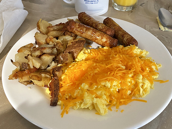 The breakfast trinity: golden home fries with crispy edges, sausage links with that perfect snap, and scrambled eggs wearing a crown of melted cheddar.