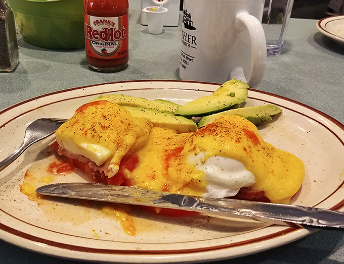 Eggs Benedict with a side of avocado&mdash;hollandaise cascading down like a golden waterfall. Frank's RedHot nearby suggests some diners like their morning with a kick.
