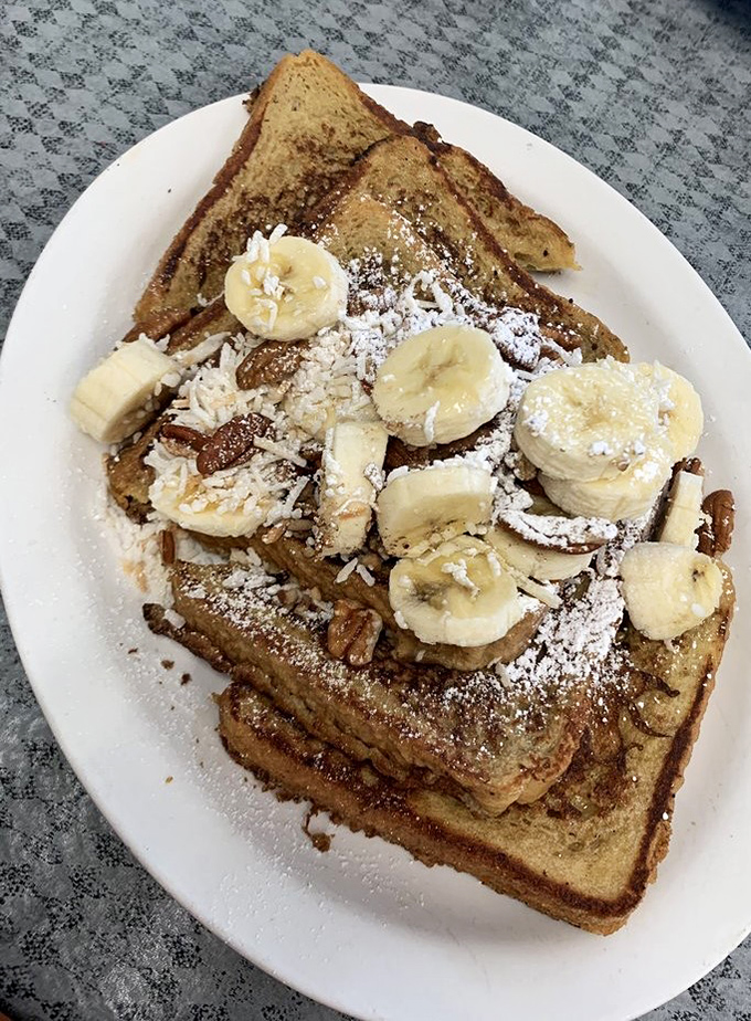 French toast that's living its best life, topped with fresh bananas, powdered sugar, and what appears to be coconut. Vacation on a plate.