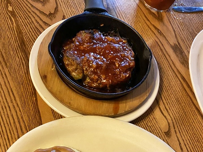 Meatloaf in its natural habitat: a sizzling skillet with sauce that glistens like ruby treasure. Comfort food royalty holding court.
