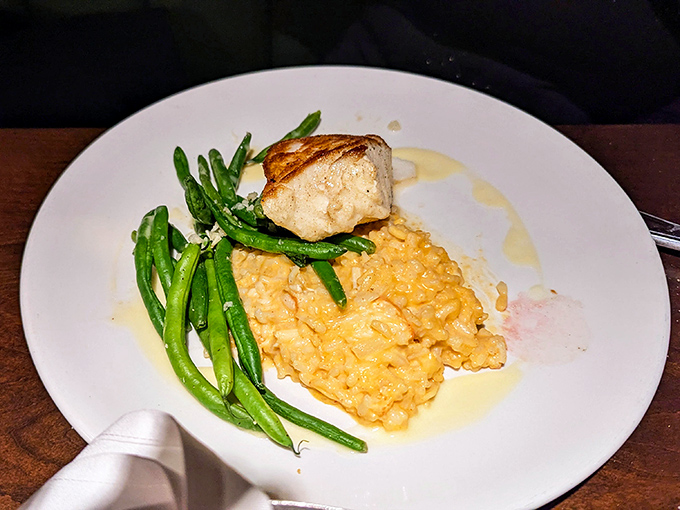 A perfectly cooked piece of fish perches atop golden risotto like a culinary island paradise, with green beans standing at attention nearby.