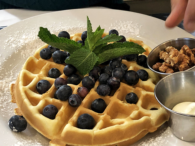 Belgian waffle perfection: golden pockets cradle fresh blueberries while mint stands guard. Those metal cups hold the secret weapons of maple and butter.