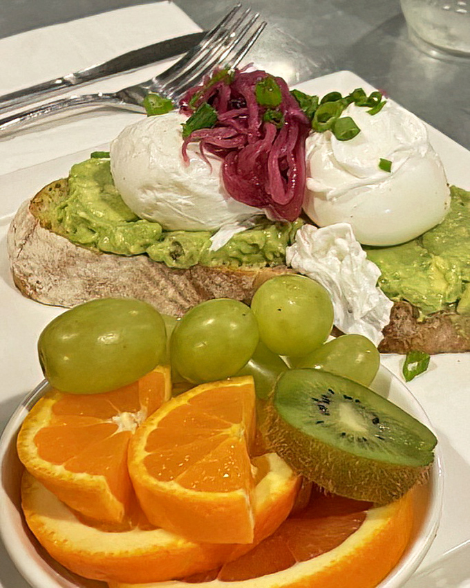 Avocado toast elevated from Instagram clich&eacute; to culinary masterpiece. Creamy avocado, perfectly poached eggs, and vibrant pickled onions atop artisanal bread with fresh fruit companions.