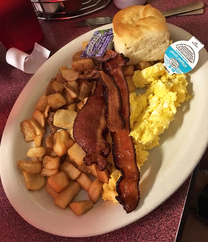 The classic breakfast trifecta: crispy bacon, fluffy scrambled eggs, and golden home fries with a biscuit standing guard. Morning salvation on a plate.