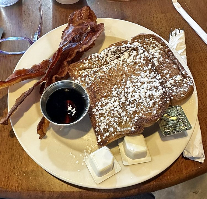 French toast dusted with powdered sugar like fresh snow, with bacon standing at attention. Maple syrup waits patiently in its metal cup for the grand pour.