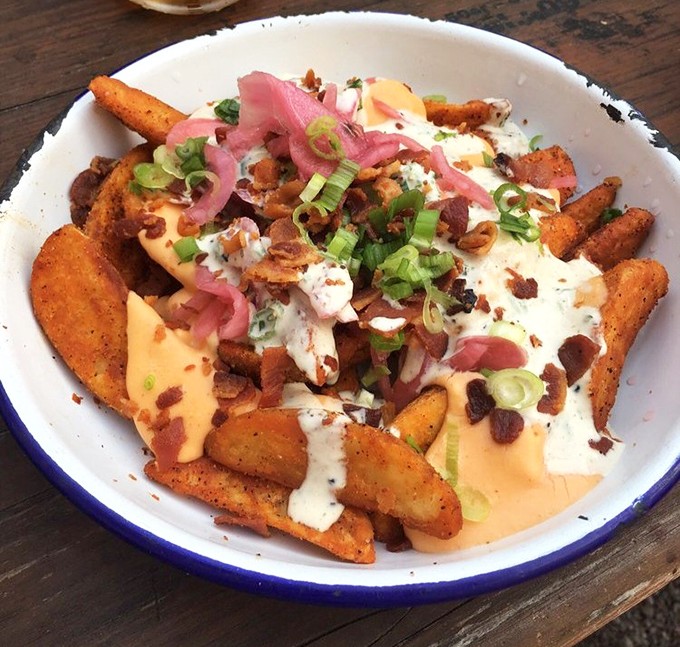 Fries living their best life under a blanket of cheese, bacon, and pickled onions. Potato paradise found.